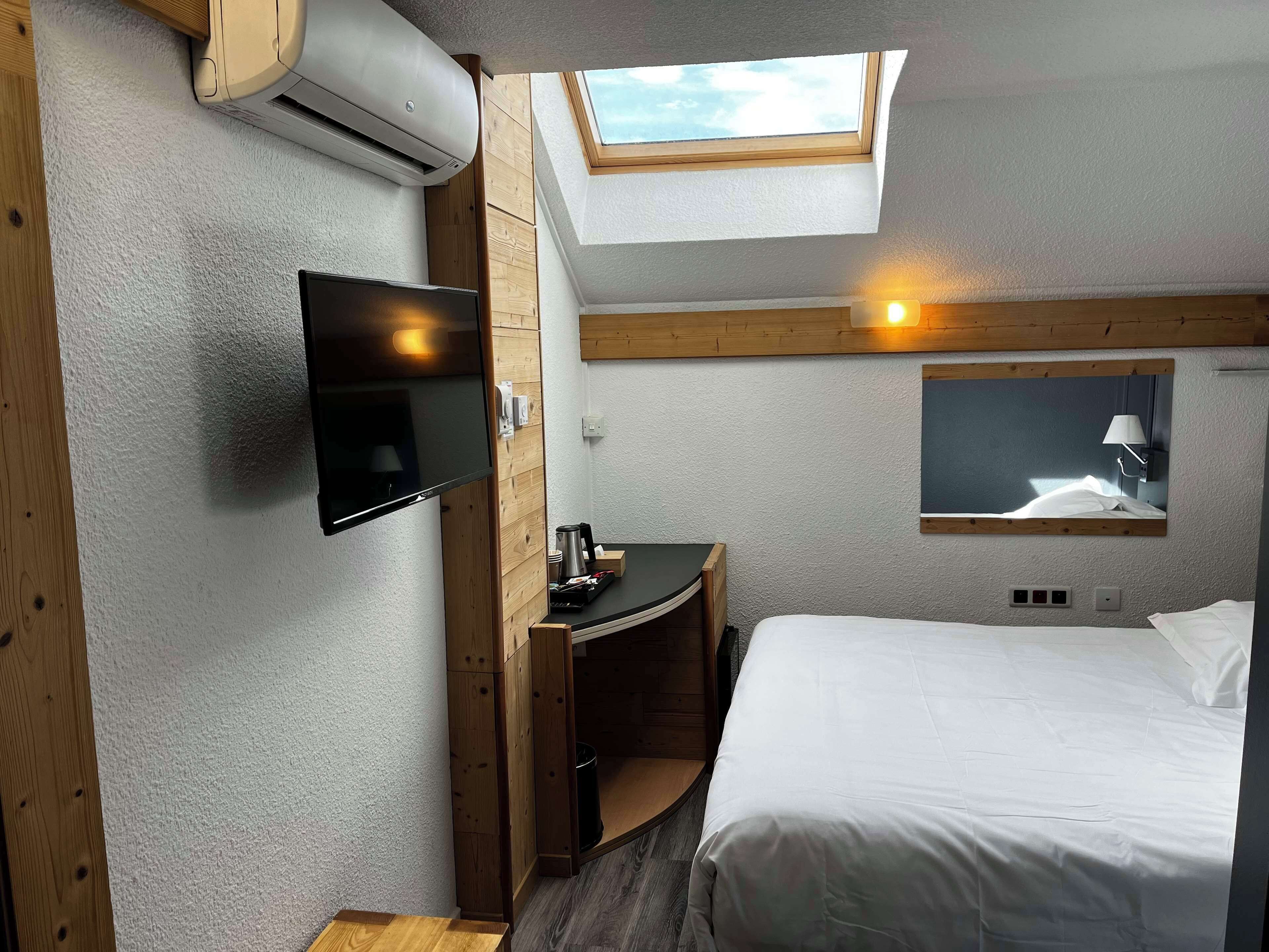 Quarto de hotel compacto com claraboia, uma cama, TV, escrivaninha com luminária e uma pequena janela.