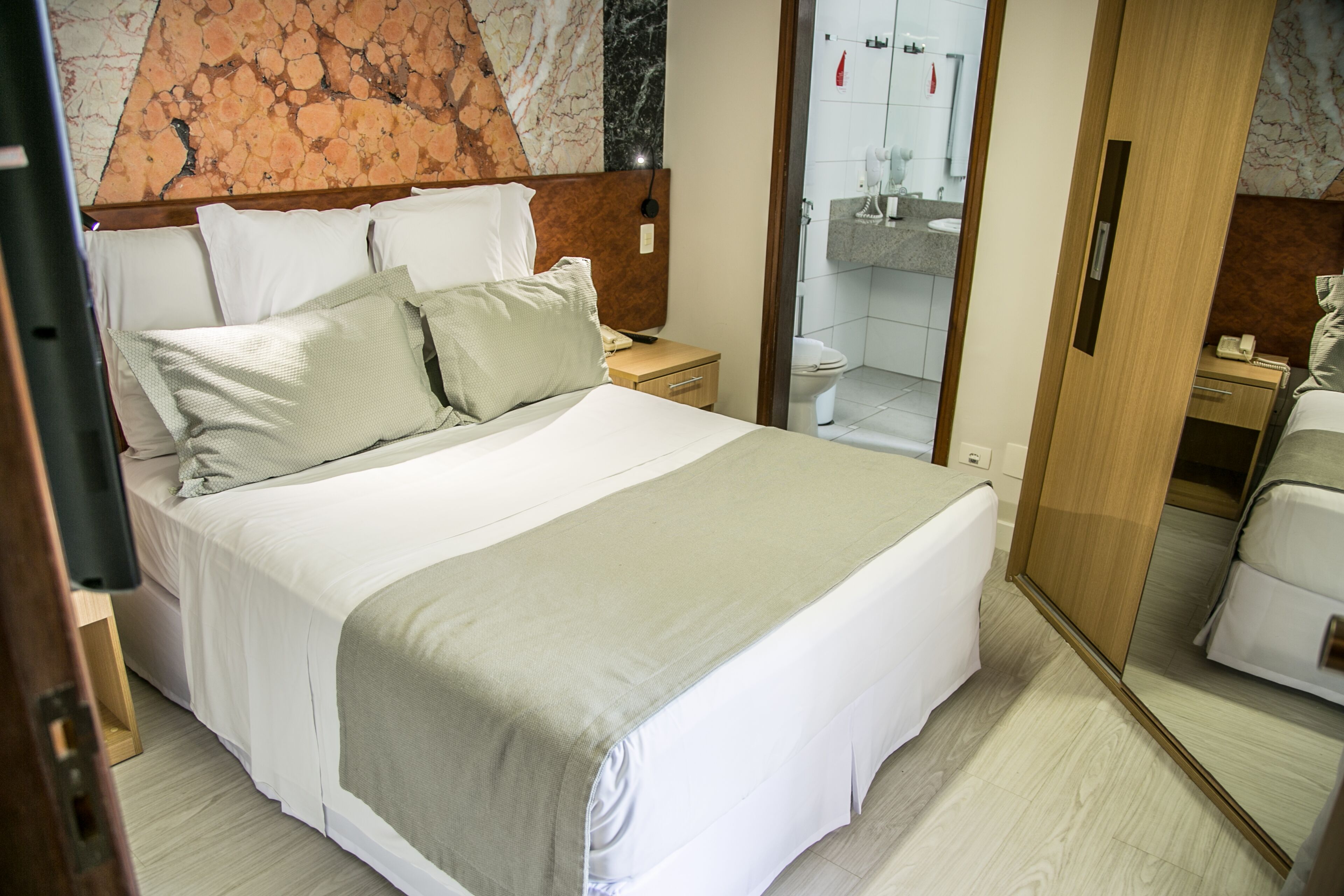 Quarto de hotel com uma cama grande, cabeceira de madeira, mesa de cabeceira, banheiro com pia e vaso sanitário, e um espelho.