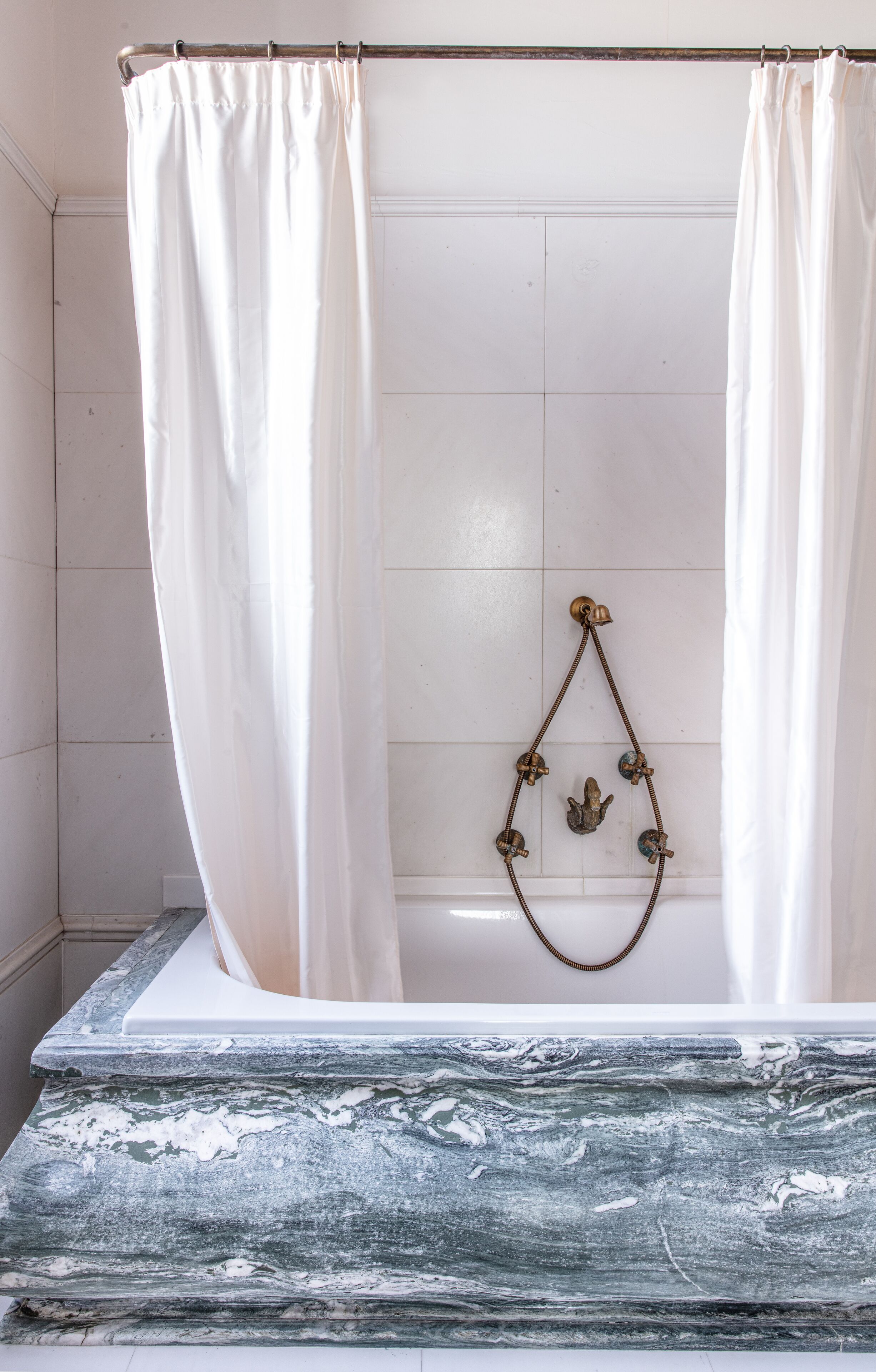 Um banheiro com banheira, cortinas de chuveiro brancas e um chuveiro com detalhe metálico decorativo.