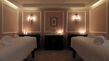Quarto de hotel com duas camas, uma mesa central e arandelas na parede.