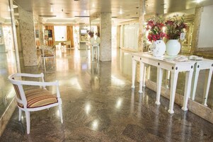 Um saguão de hotel com piso de mármore, balcão de recepção, uma cadeira branca com almofada listrada e um vaso com flores.