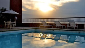 Piscina com espreguiçadeiras e guarda-sóis, refletindo o pôr do sol.