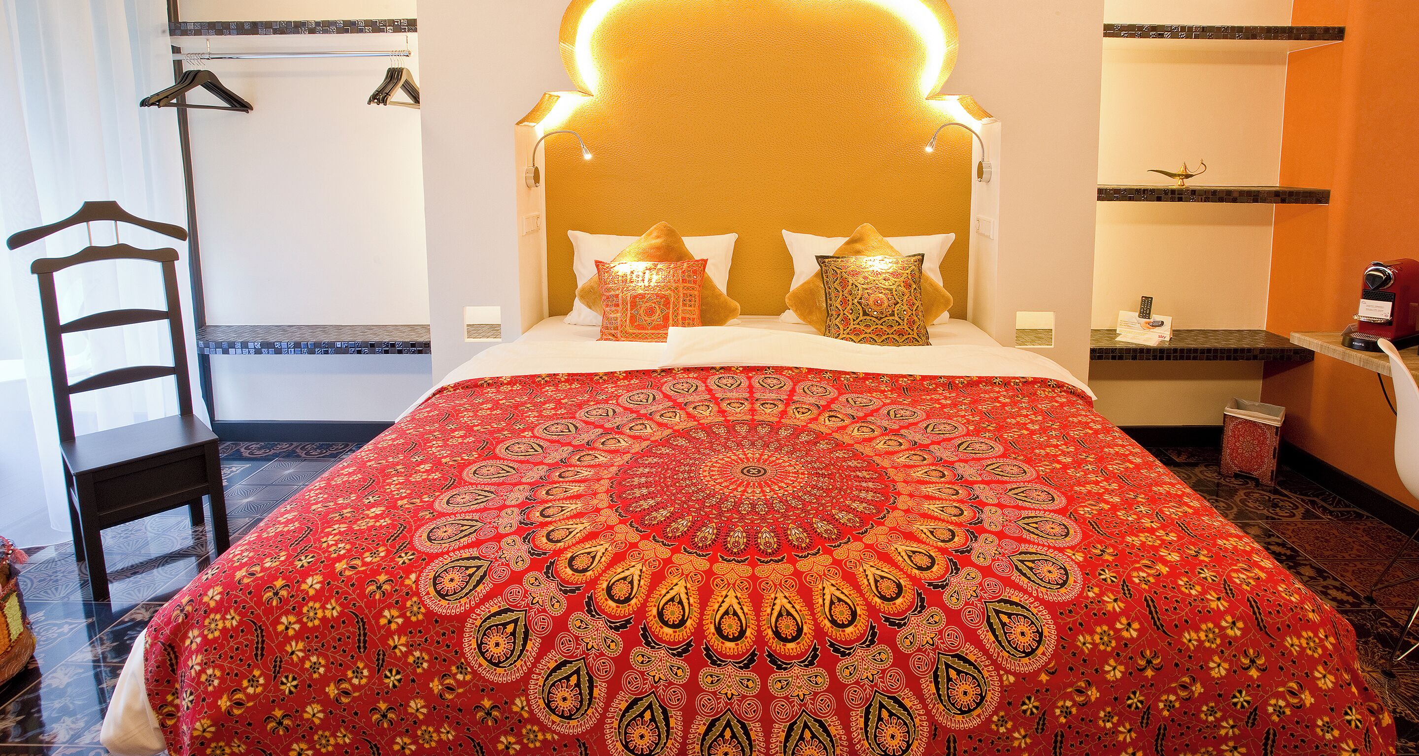 Quarto de hotel com uma cama grande, cabeceira com iluminação integrada, mesa de cabeceira com abajur e uma pequena prateleira com objetos decorativos.