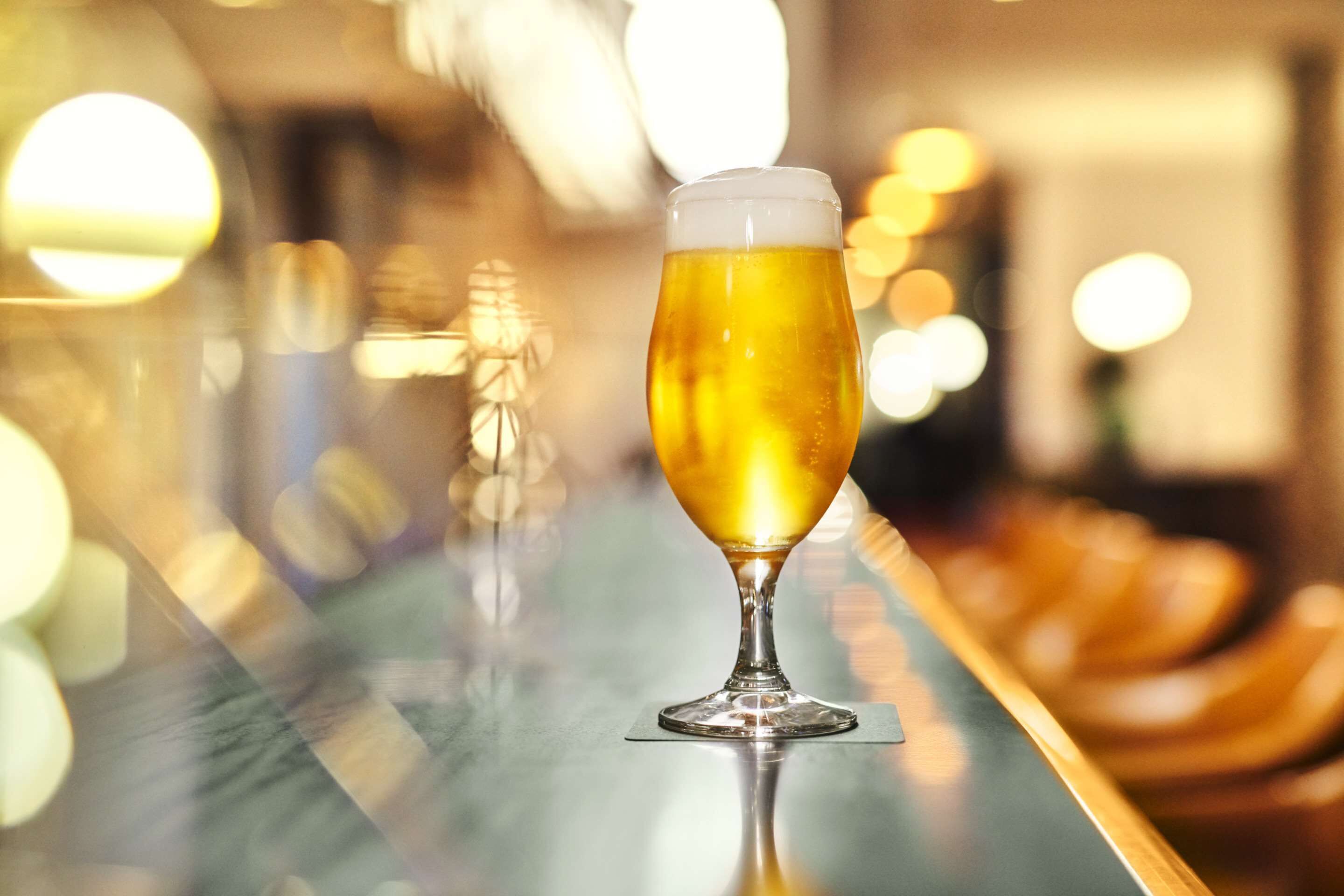 Um copo de cerveja sobre o balcão de um bar, com luzes e assentos ao fundo desfocados.