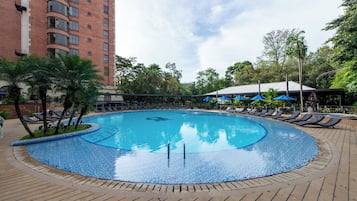 Piscina com borda em azulejo, rodeada por espreguiçadeiras e guarda-sóis, com um edifício alto ao fundo.
