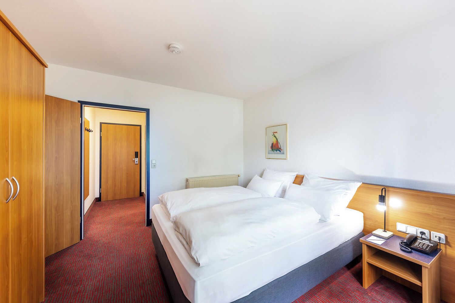 Quarto de hotel com uma cama, mesa de cabeceira com abajur, telefone e porta que dá para outro quarto.