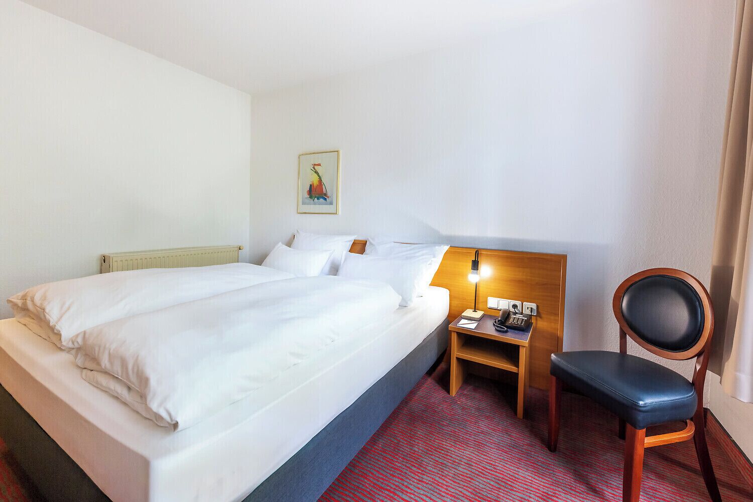 Quarto de hotel com cama, mesa de cabeceira, abajur, cadeira e um quadro na parede.