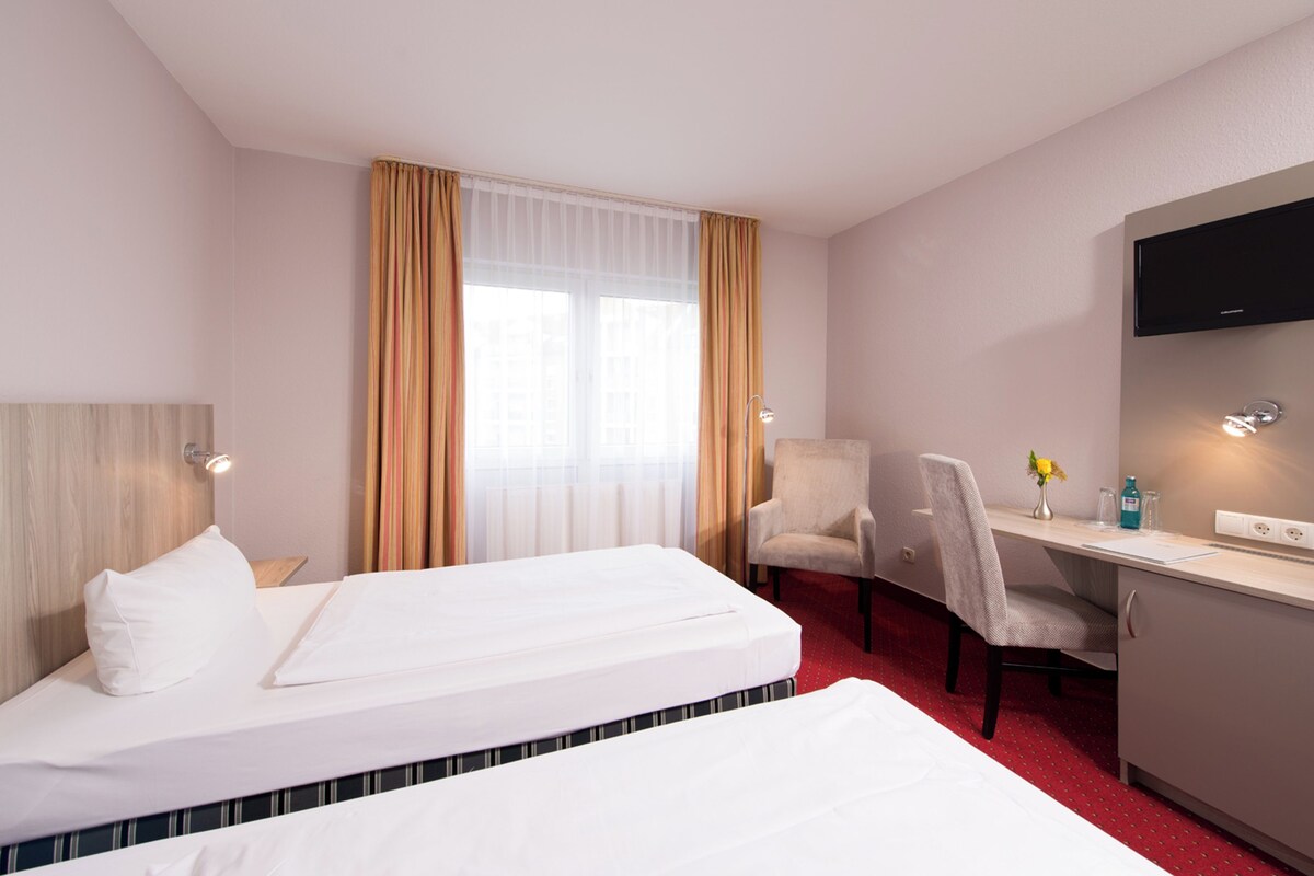 Quarto de hotel com cama, escrivaninha, cadeira e televisão.