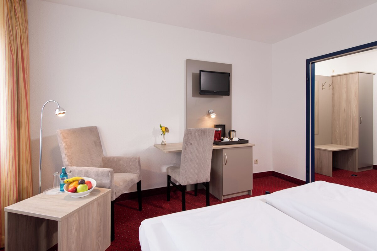 Quarto de hotel com cama, escrivaninha e cadeira, televisão na parede, mesa com fruteira e espelho.