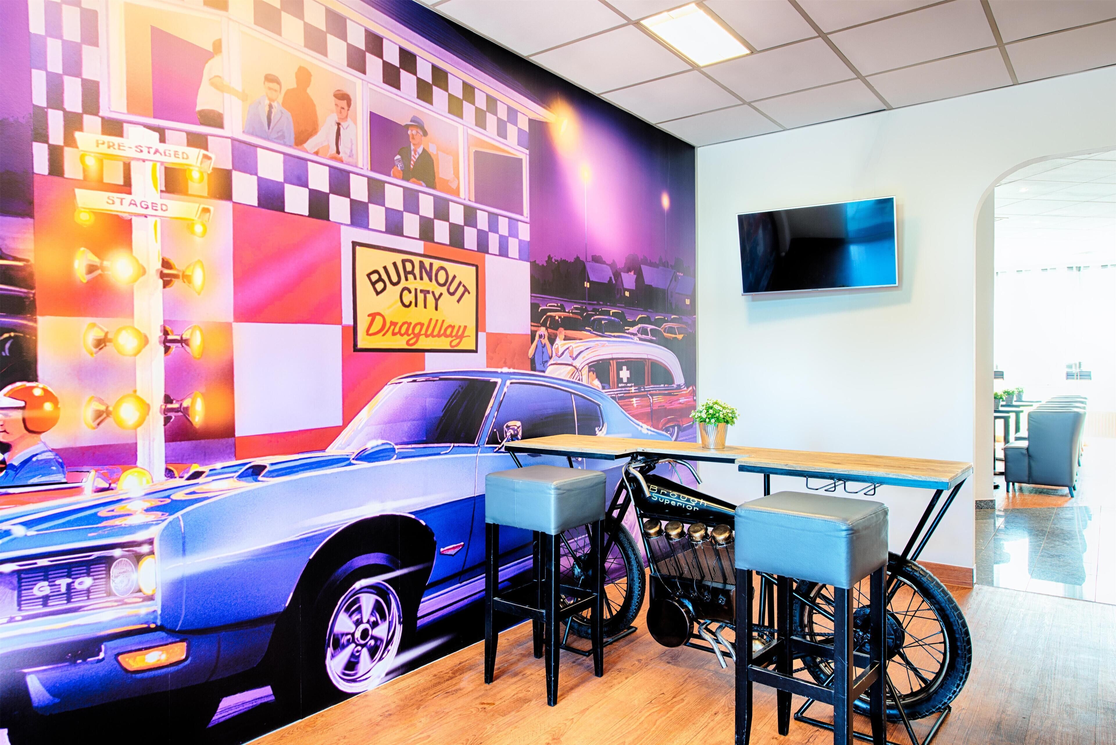 Quarto com um mural de carro de corrida de arrancada, área de bar com banquetas e uma televisão na parede.