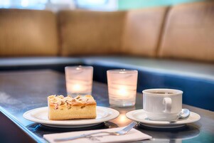 Uma fatia de bolo num prato, uma chávena de café e duas velas sobre uma mesa.
