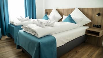 Quarto de hotel com duas camas, cada uma com roupa de cama azul turquesa e almofadas brancas. Apresenta uma cabeceira de madeira, uma mesa de cabeceira com abajur e piso de madeira.