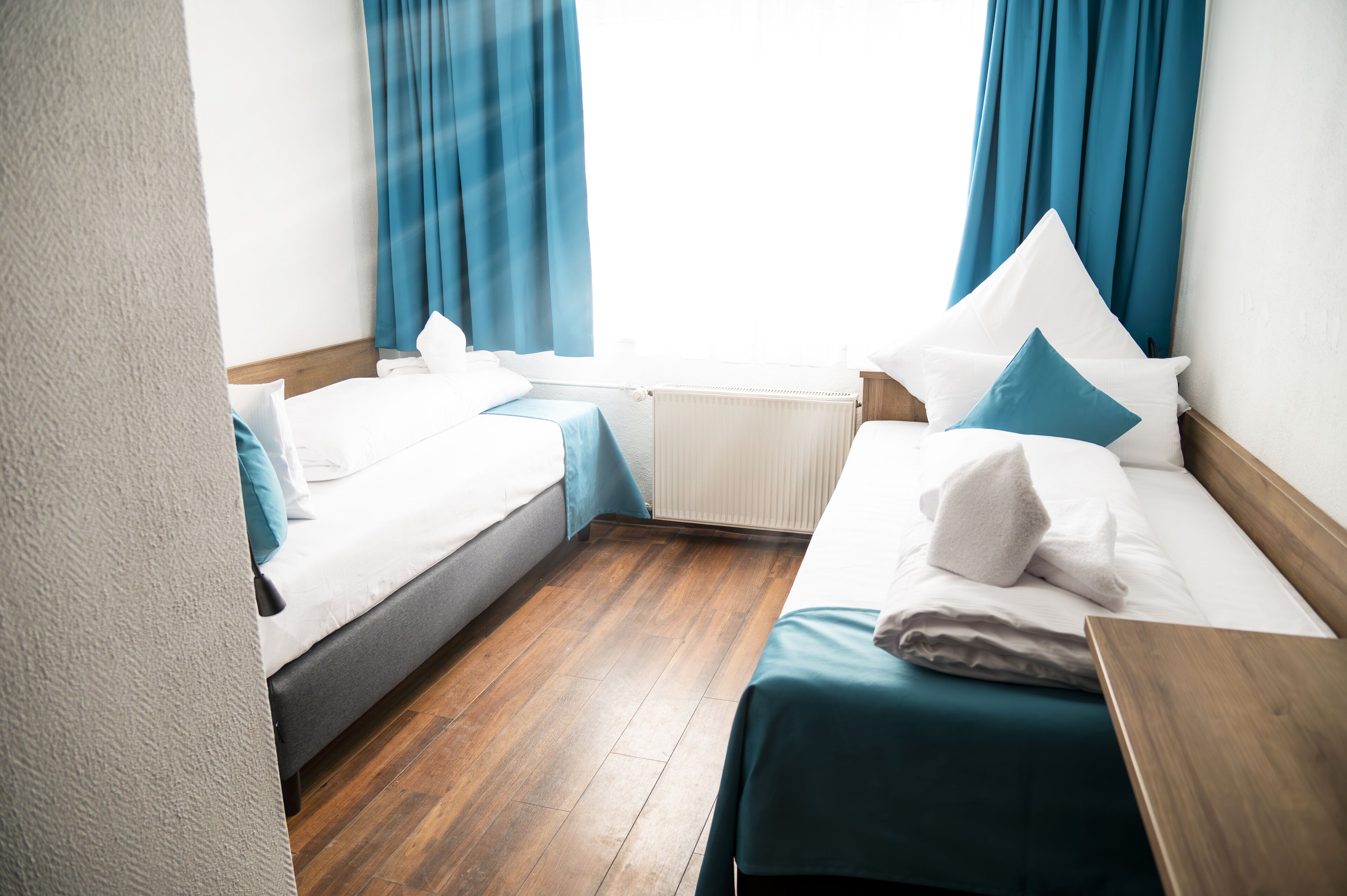 Quarto de hotel com duas camas de solteiro, piso de madeira, radiador e cortinas azuis.