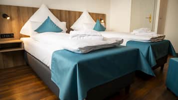Quarto de hotel com duas camas de solteiro, cada uma com colcha verde-água e lençóis brancos. Há uma cabeceira de madeira, uma mesa de cabeceira com abajur e uma porta com puxador dourado.