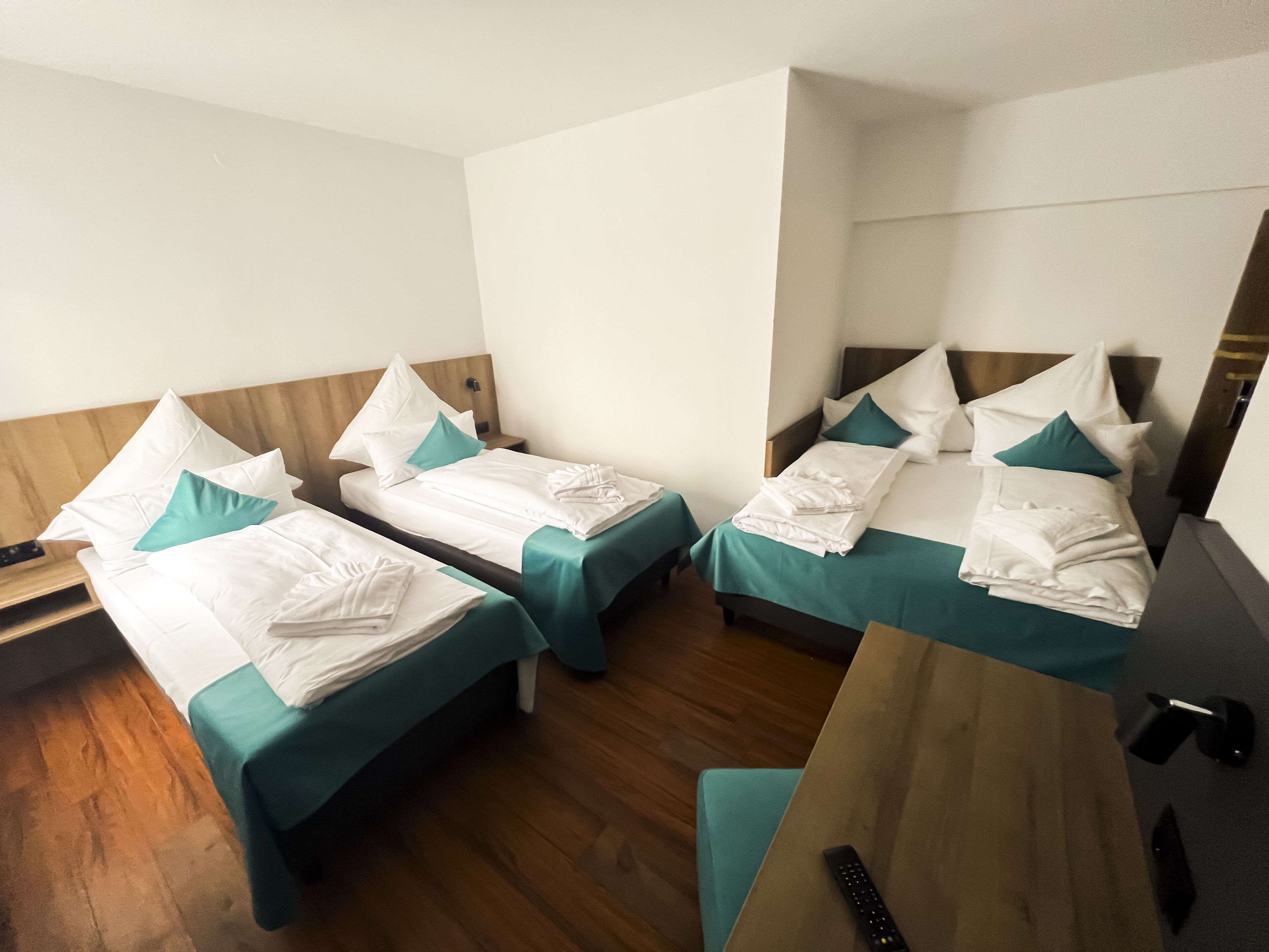 Quarto de hotel com duas camas de solteiro, cada uma com cabeceira de madeira, roupa de cama branca e detalhes em verde-água. Há uma escrivaninha de madeira com um controle remoto.