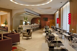 Um saguão moderno de hotel com bar, áreas de estar e uma grande janela com detalhes decorativos vermelhos.