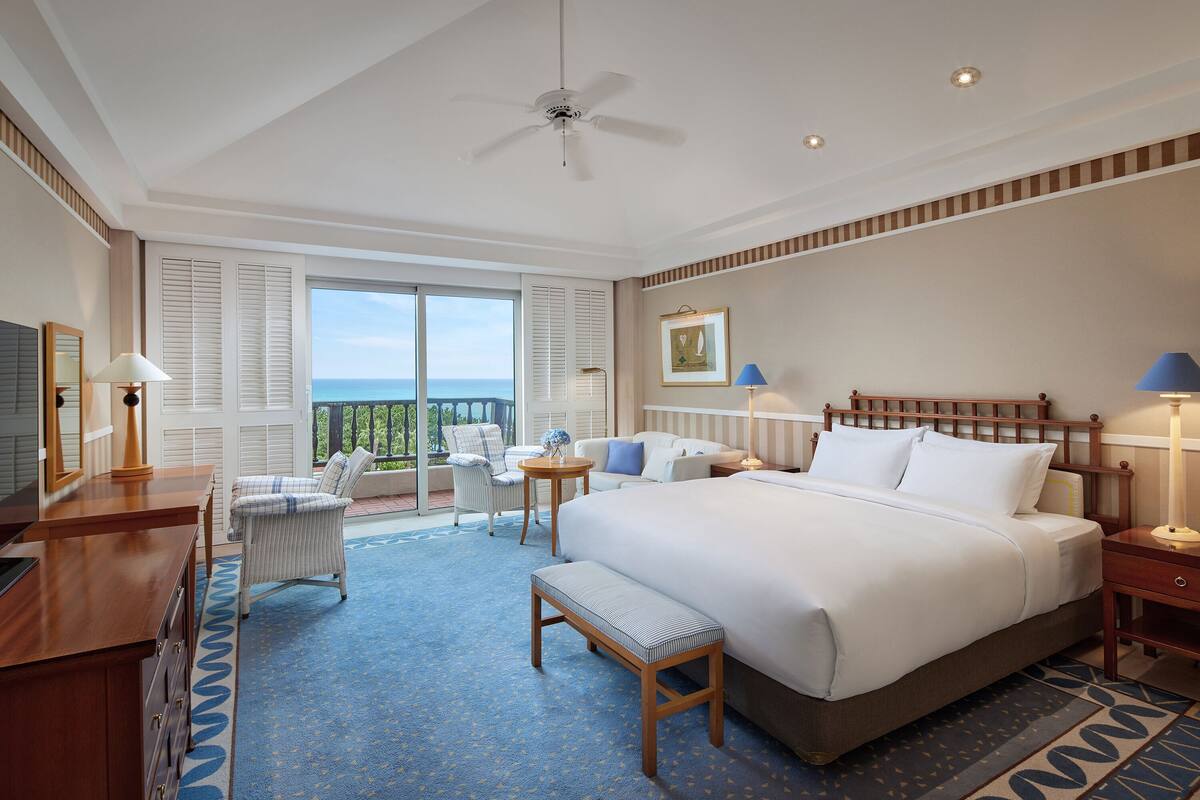 Quarto de hotel com uma cama grande, uma escrivaninha, uma cadeira, uma mesinha e varanda com vista.