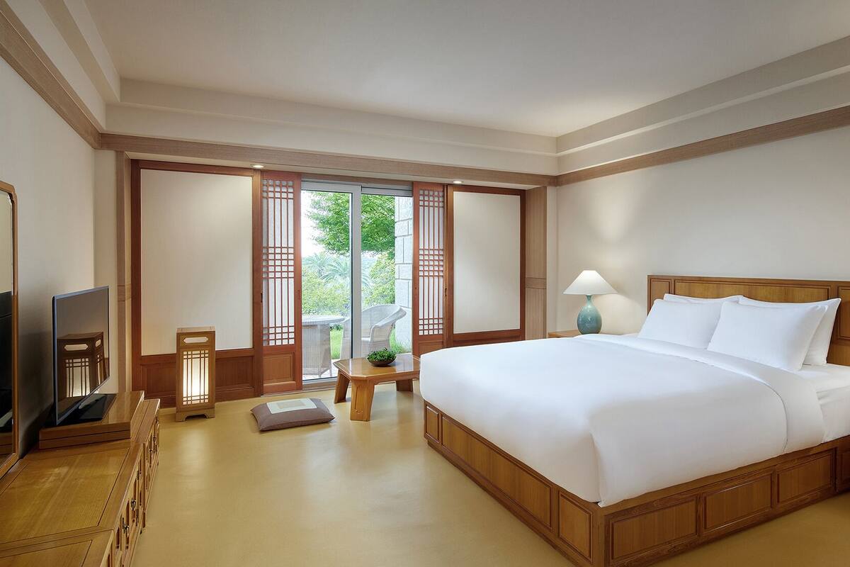 Quarto de hotel com uma cama grande, televisão, armário de madeira, pequena mesa com uma planta e porta de correr para uma varanda.