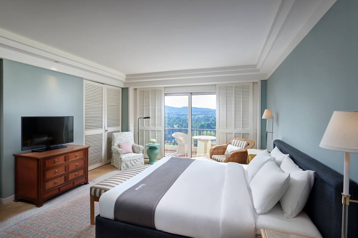 Quarto de hotel com uma cama grande, televisão, penteadeira, varanda com vista para as montanhas e área de refeições com mesa e cadeiras.