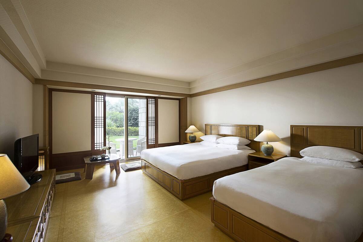 Quarto de hotel com duas camas, televisão, escrivaninha e janela com vista para o verde.