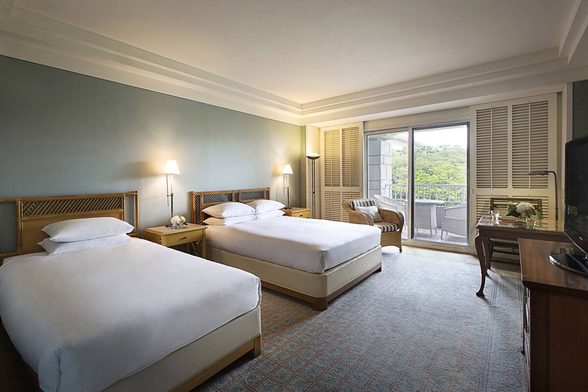Quarto de hotel com duas camas, televisão, escrivaninha e varanda com vista para o verde.