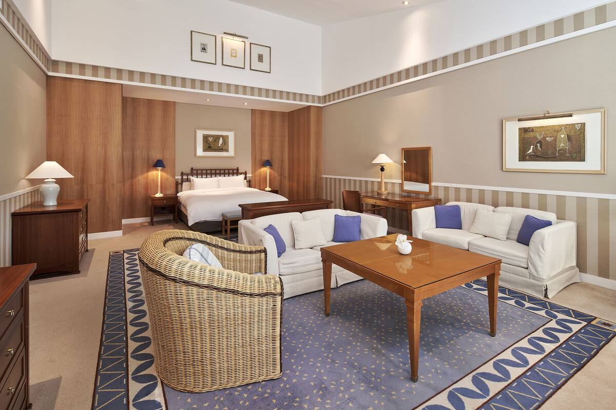 Quarto de hotel com cama, dois sofás brancos, uma poltrona de vime, uma mesa de madeira e um tapete estampado.