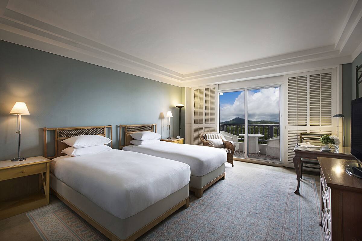 Quarto de hotel com duas camas, varanda com vista, escrivaninha com televisão e uma cadeira.