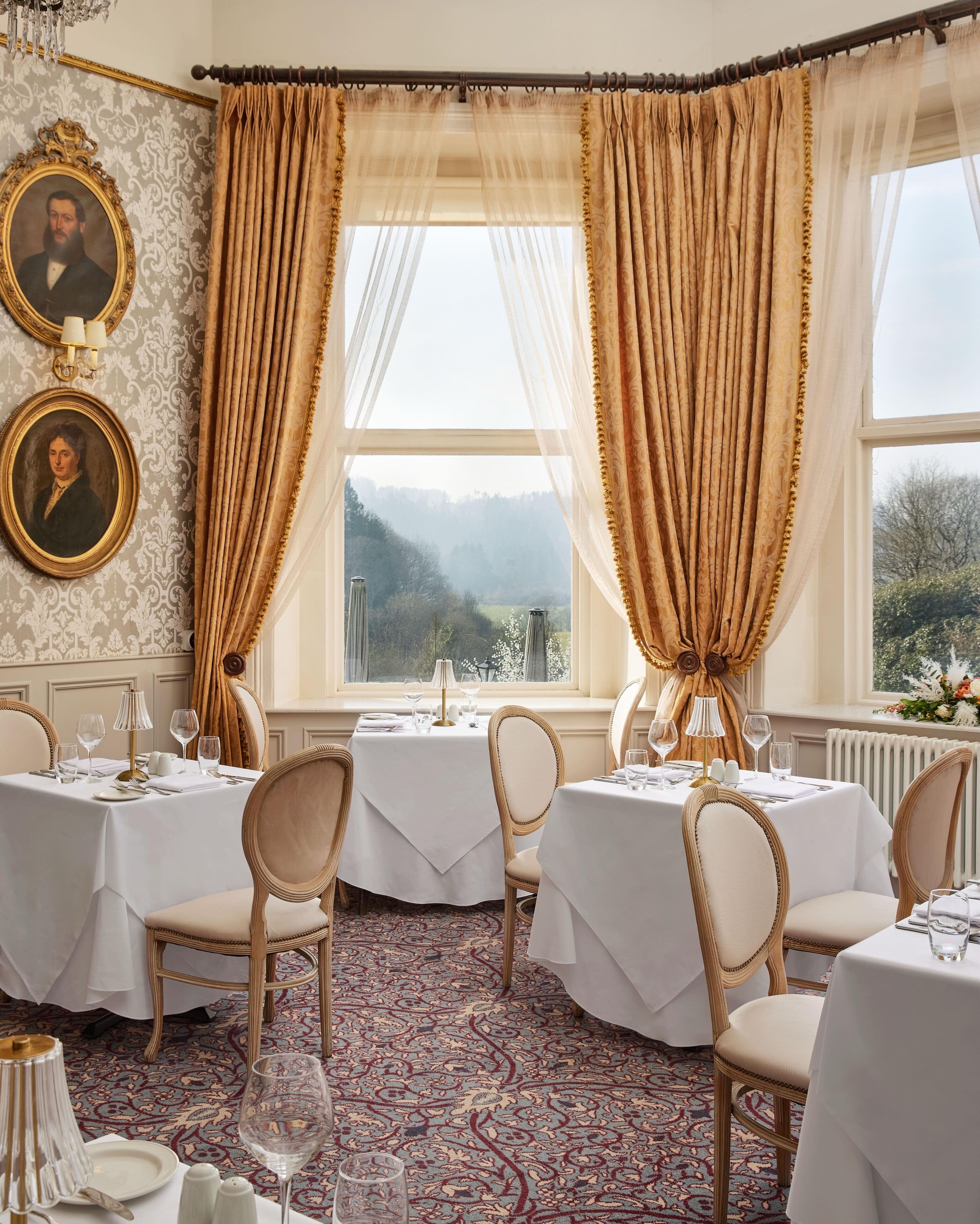 Uma sala de jantar com quatro mesas, cada uma com toalhas brancas, cadeiras e copos. O espaço apresenta papel de parede trabalhado, um candelabro e grandes janelas com cortinas.