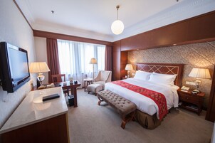 Quarto de hotel com uma cama grande, uma escrivaninha com televisão, uma cadeira, uma mesa pequena e um banco.