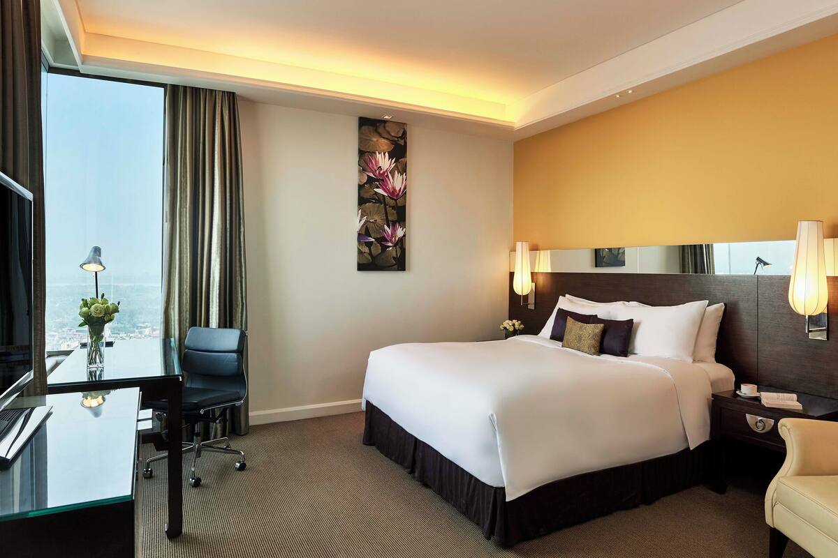 Quarto de hotel com uma cama grande, uma escrivaninha com cadeira, uma luminária de mesa, uma mesa de cabeceira, uma poltrona e um quadro na parede.