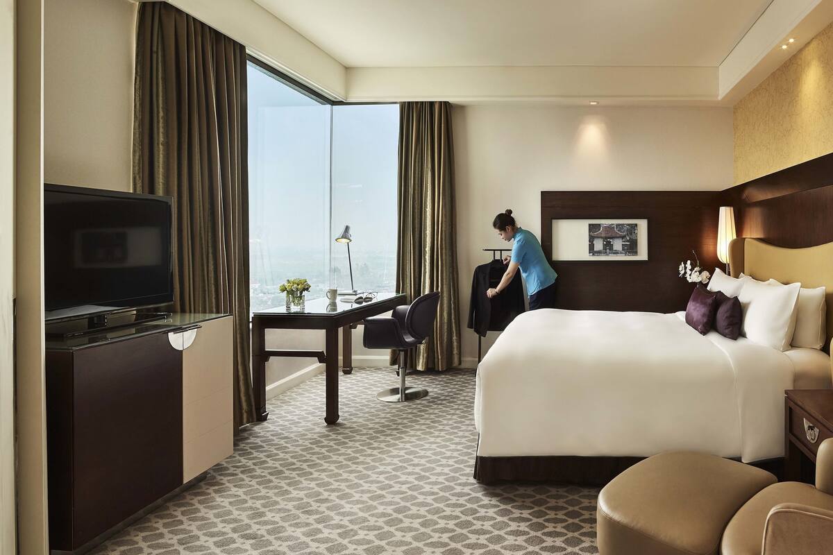 Quarto de hotel com uma cama grande, uma escrivaninha com cadeira, televisão e vista para a cidade através de grandes janelas.