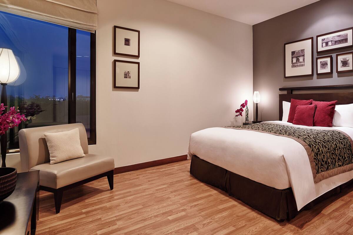 Quarto de hotel com cama, uma cadeira, escrivaninha, vaso de flores e quadros nas paredes.