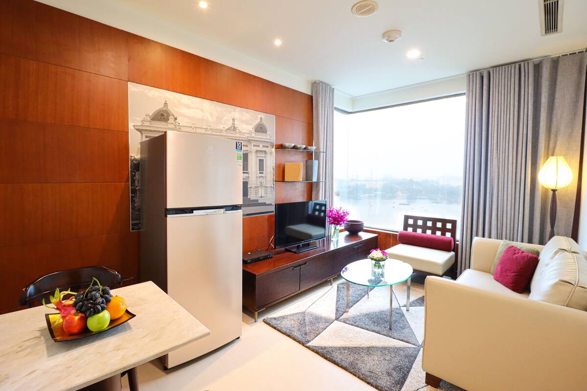 Quarto de hotel moderno com área de refeições, sofá, TV e uma grande janela com vista para a cidade.