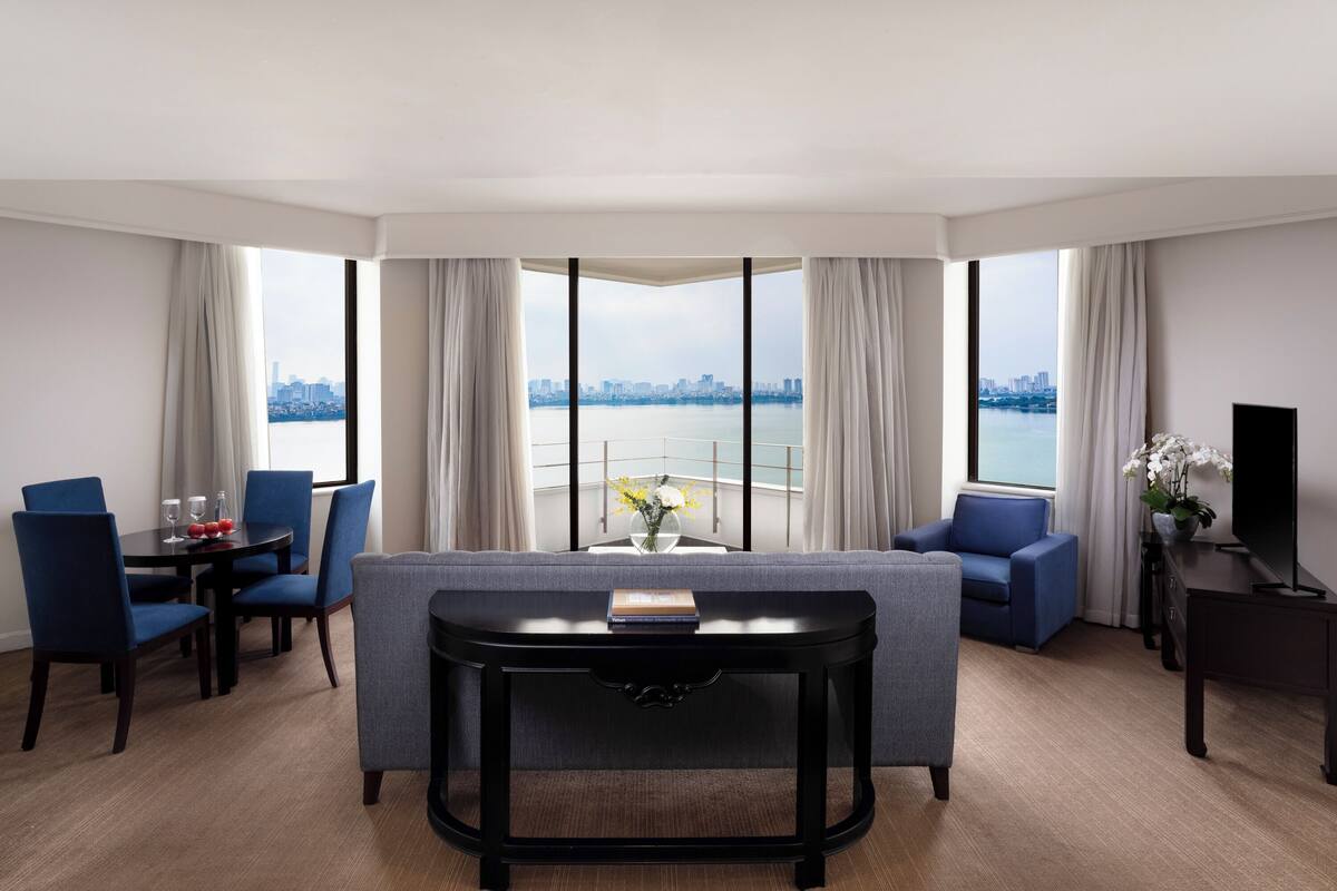 Quarto de hotel moderno com uma janela grande dando vista para a cidade, um sofá, uma mesa de jantar e um rack para televisão.