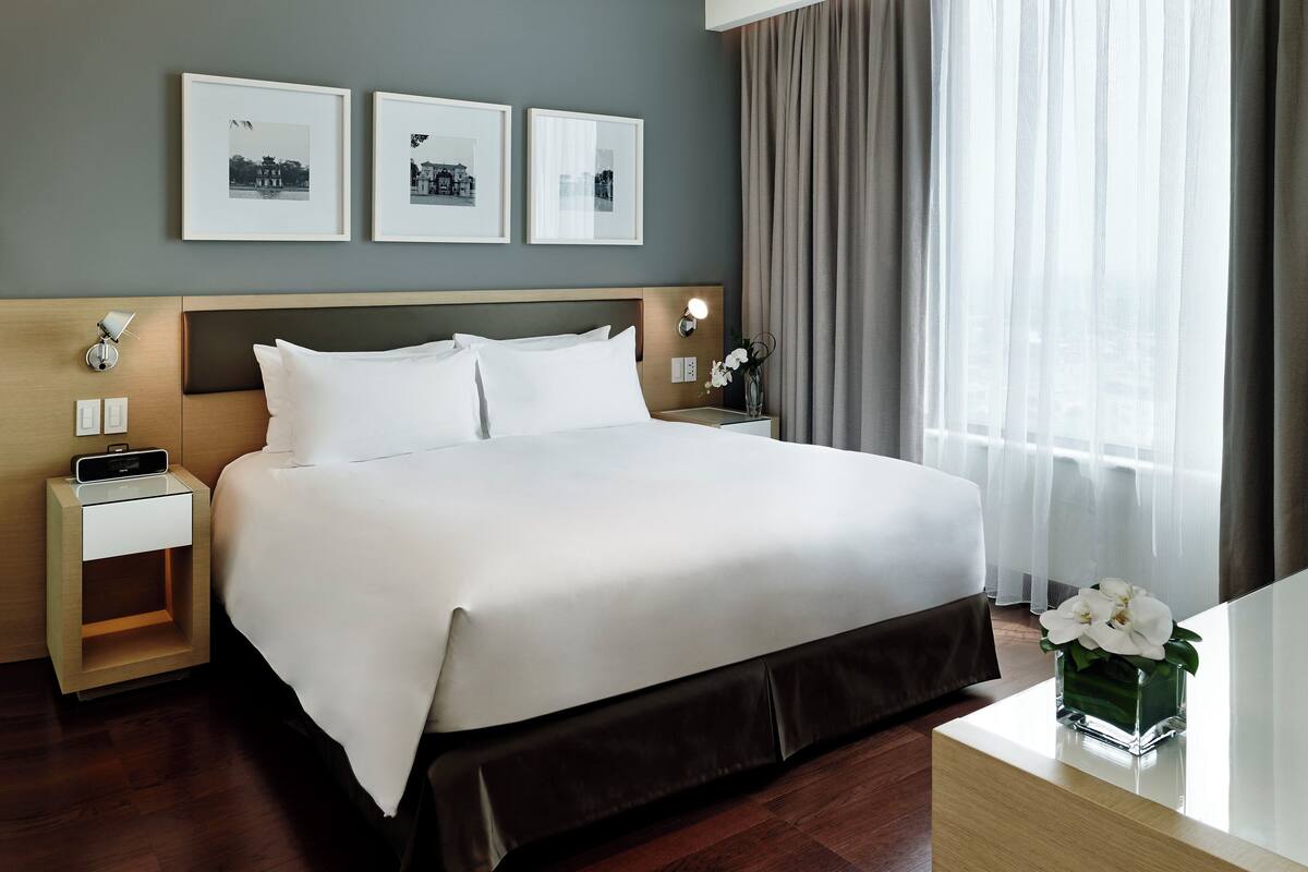Quarto de hotel bem arrumado com uma cama grande, uma mesa de cabeceira com abajur, um criado-mudo com telefone e uma janela com cortinas.