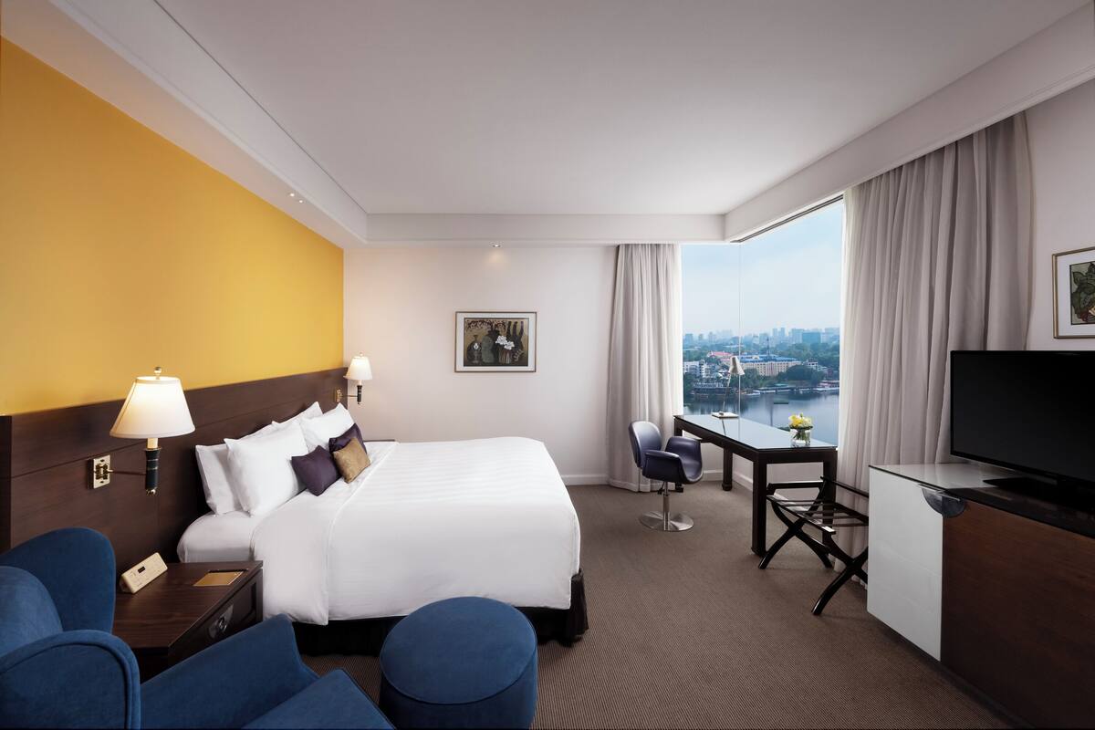 Quarto de hotel com uma cama grande, uma mesa, uma cadeira, uma TV e vista para a paisagem urbana.