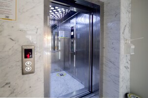 Interior de elevador com paredes de mármore, painel de comando com botões de direção e chão com sinal de alerta circular.
