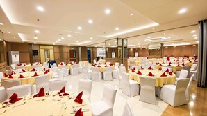 Salão de banquetes com mesas redondas montadas para refeições, cadeiras brancas e guardanapos vermelhos.