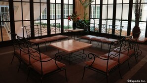 Um café com uma mesa central rodeada por cadeiras estofadas, janelas amplas e plantas em vasos.
