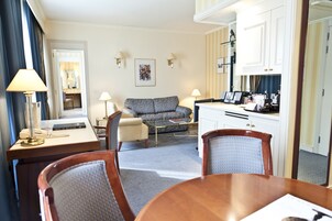 Quarto de hotel com mesa de jantar, cadeiras, sofá, escrivaninha com luminária e cozinha compacta.