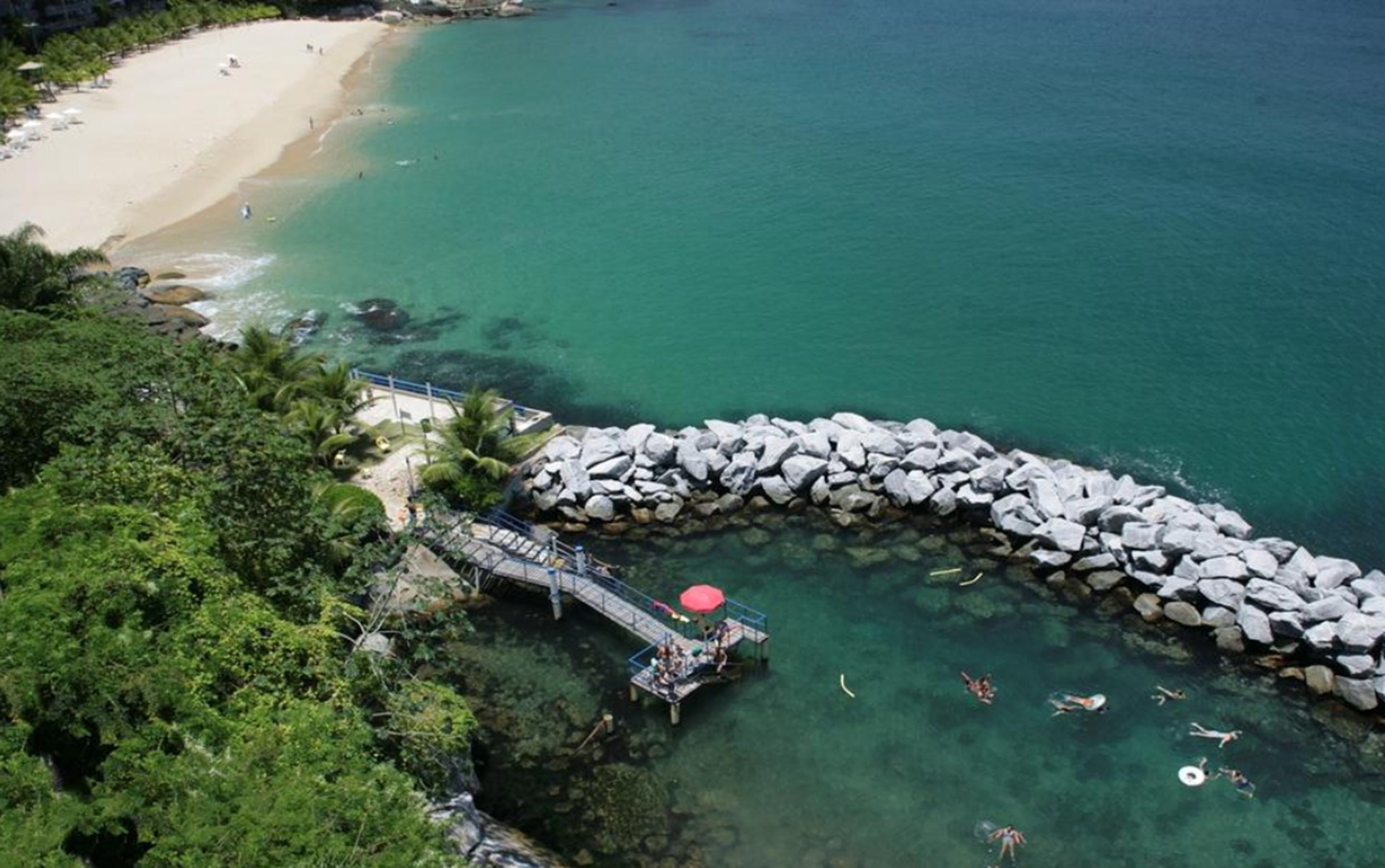 Vista aérea de uma praia com águas turquesas cristalinas, faixa de areia e um quebra-mar rochoso.