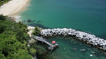 Vista aérea de uma praia com águas turquesas cristalinas, faixa de areia e um quebra-mar rochoso.