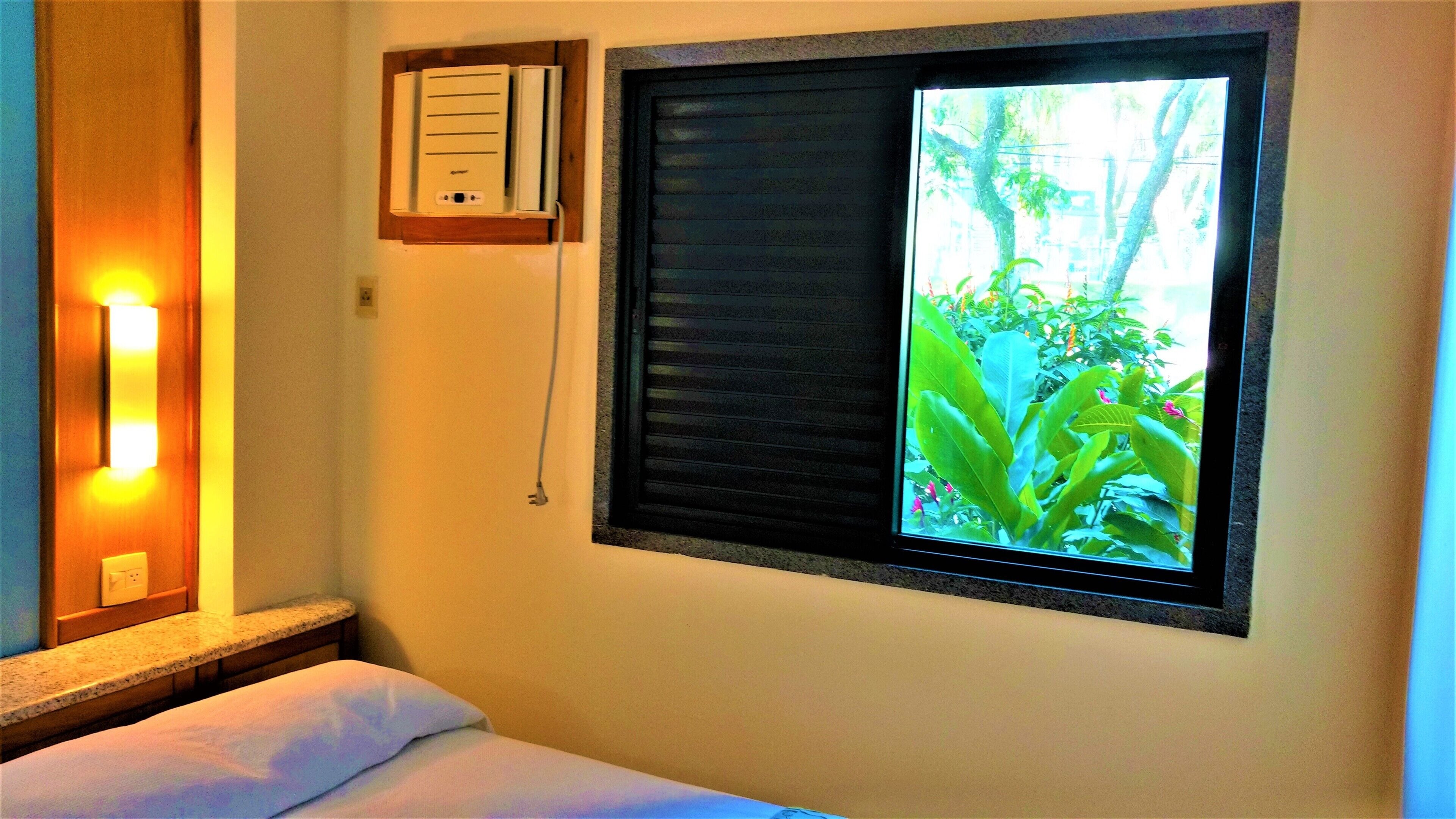 Quarto de hotel com cama, ar condicionado fixado na parede e janela com vista para áreas verdes.