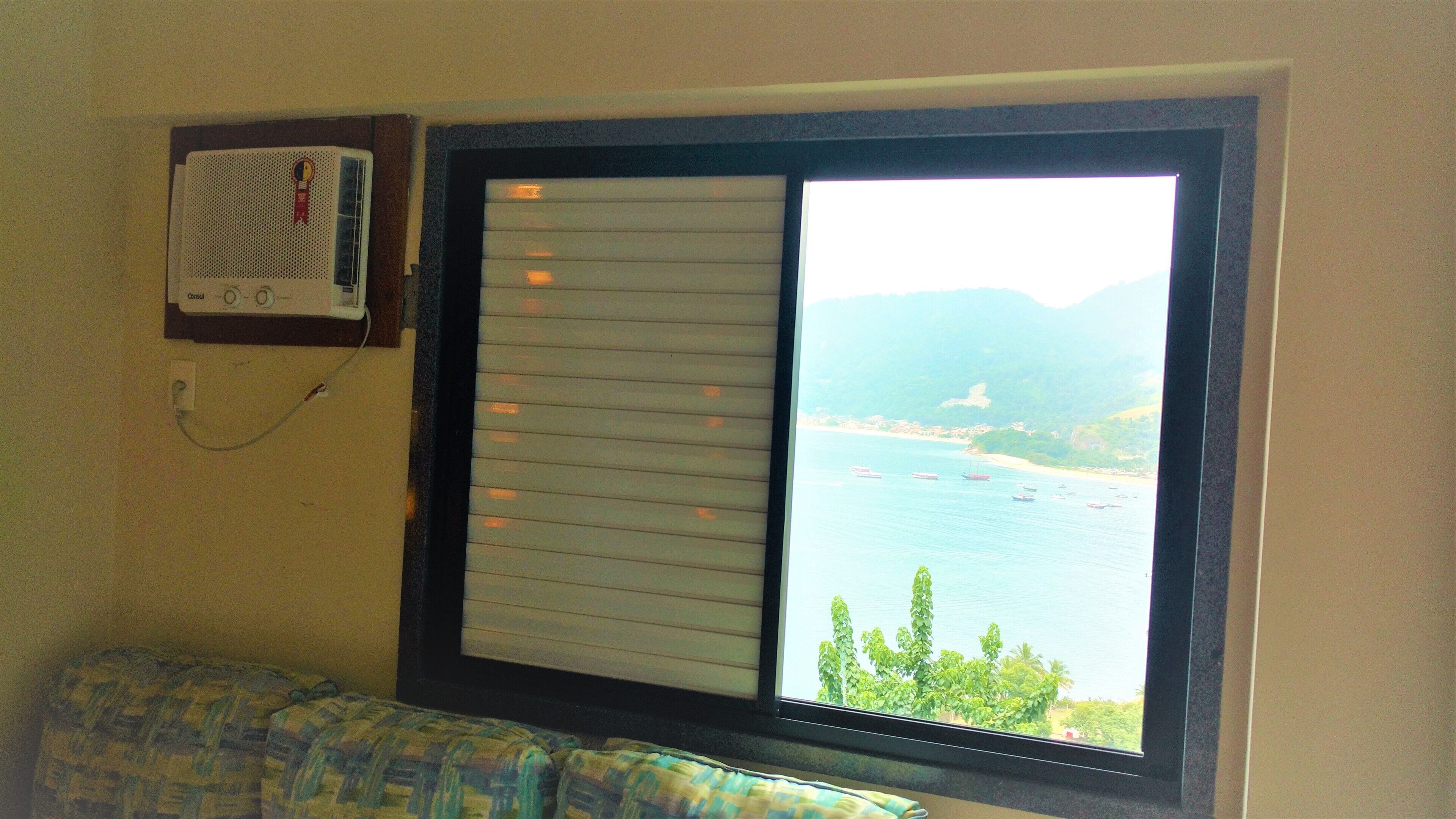 Quarto com janela e vista para o litoral, cama com roupa de cama estampada e ar condicionado fixado na parede.