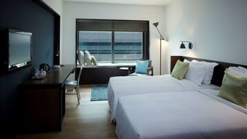 Quarto de hotel com cama, escrivaninha com cadeira, televisão e janela com vista.