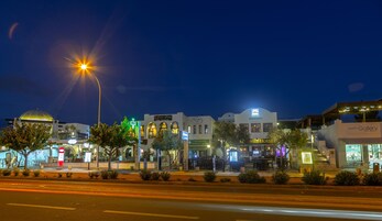 Vista da rua à noite, com edifícios iluminados, incluindo uma farmácia, restaurante e galeria.