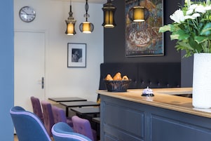 Interior de restaurante com balcão de madeira, cadeiras roxas e azuis, uma cesta de croissants e um vaso branco grande com flores.