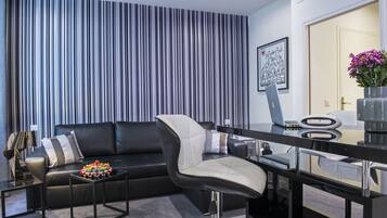 Uma sala de estar moderna com um sofá de couro preto, uma mesinha lateral com um objeto colorido, uma poltrona preta e branca, uma mesa de centro de vidro e uma parede listrada.