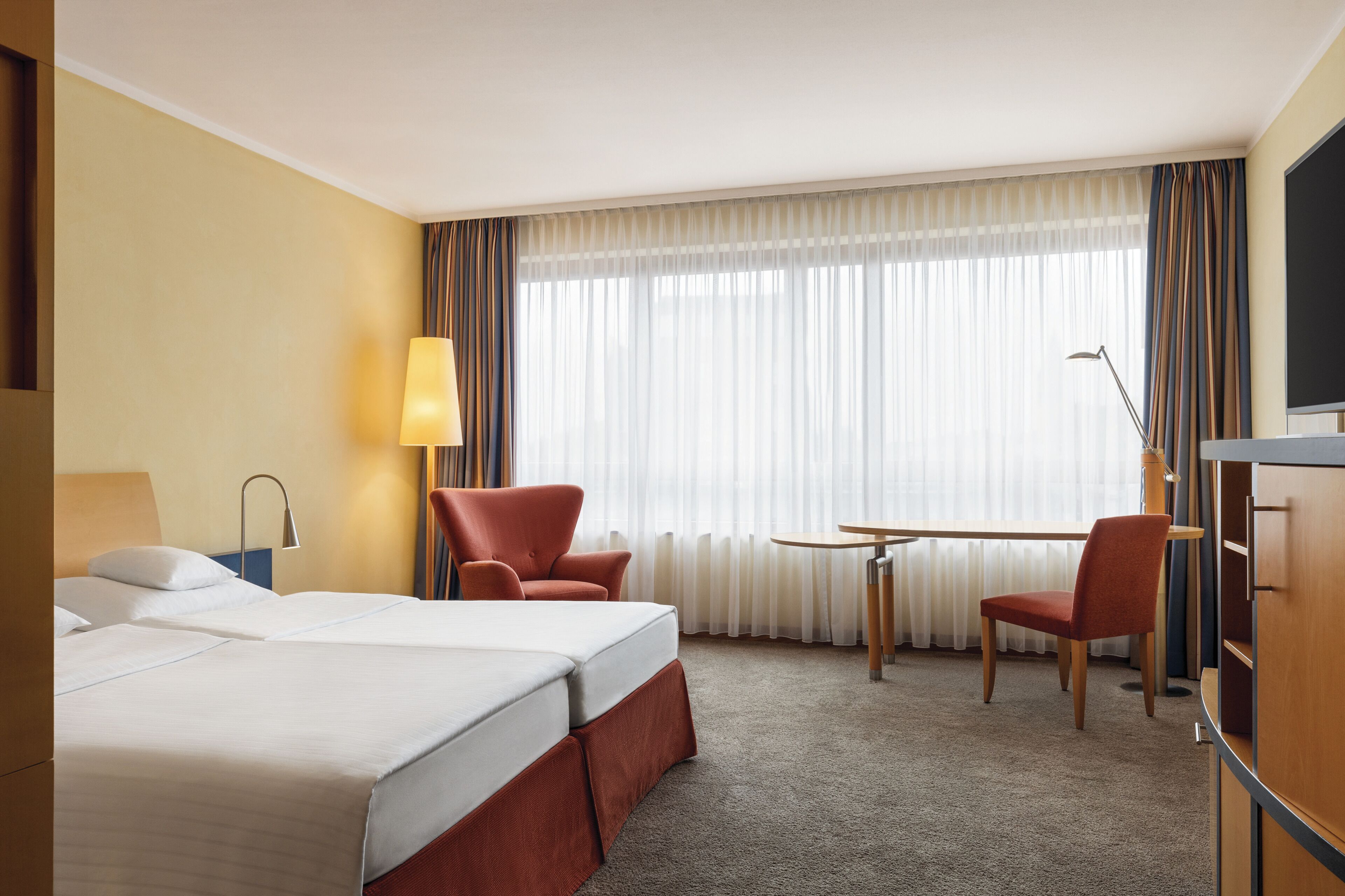 Quarto de hotel com cama, duas cadeiras, uma mesa, abajur e televisão.