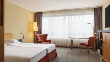 Quarto de hotel com cama, duas cadeiras, uma mesa, abajur e televisão.
